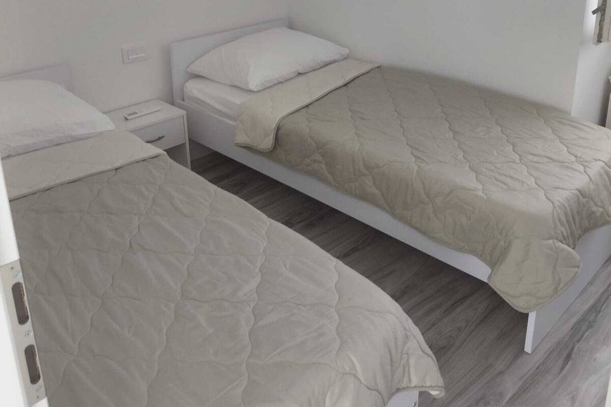Um quarto pequeno e limpo, com duas camas de solteiro, uma mesa de cabeceira e uma parede branca.