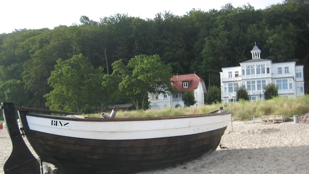 Um barco numa praia arenosa, com uma floresta e edifícios ao fundo.