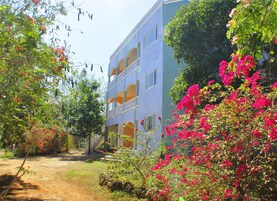 Edifício de vários andares com sacadas, cercado por vegetação exuberante e flores rosa vibrantes.