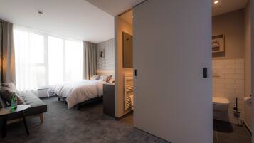 Quarto de hotel moderno com sofá, duas camas, uma pequena mesa e vista para a cidade.