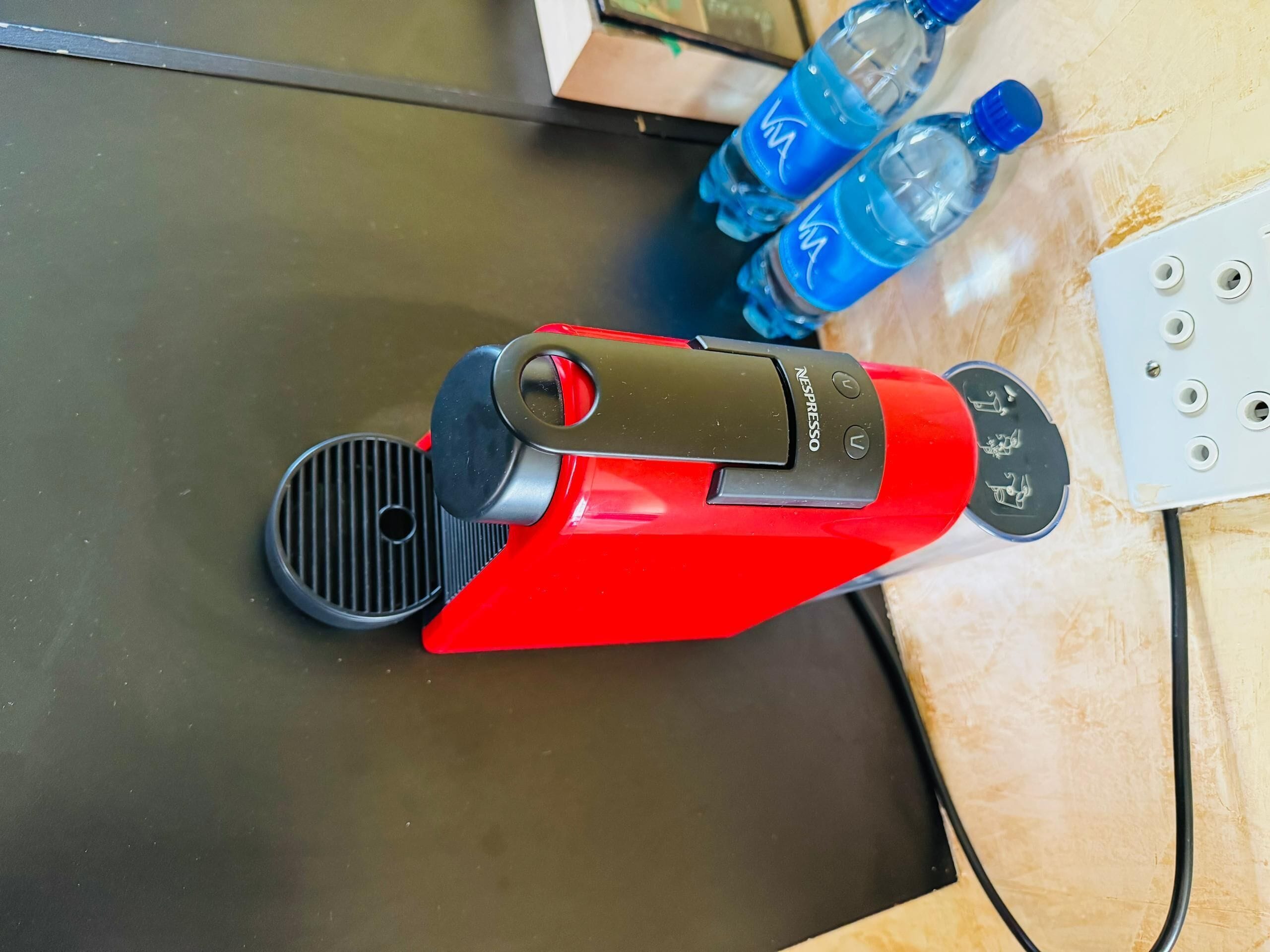 Uma máquina de café Nespresso vermelha sobre uma bancada escura, com duas garrafas de água azuis ao fundo.