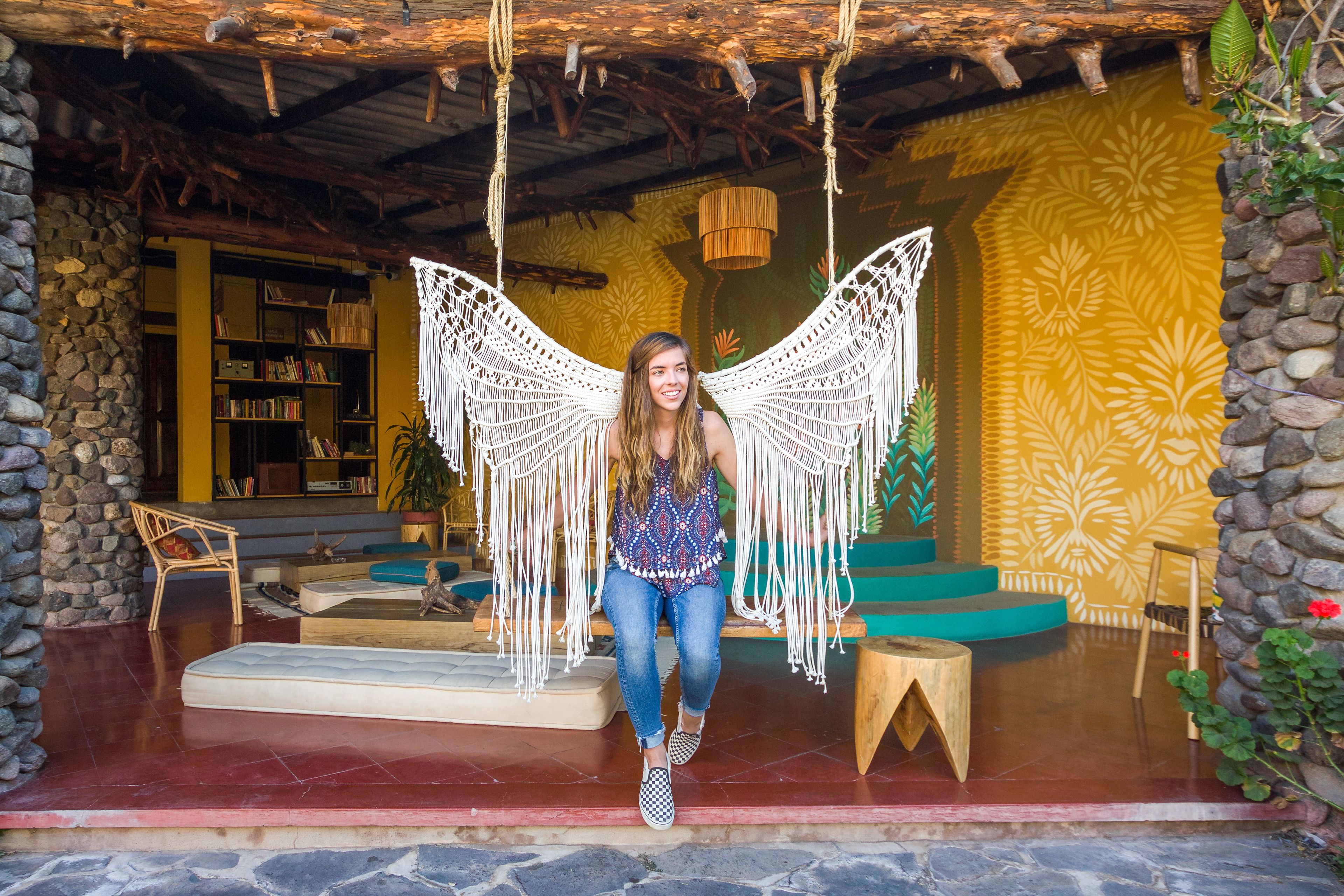 Mulher sentada num balanço com asas de macramé, parede de pedra, estante de livros e parede estampada ao fundo.