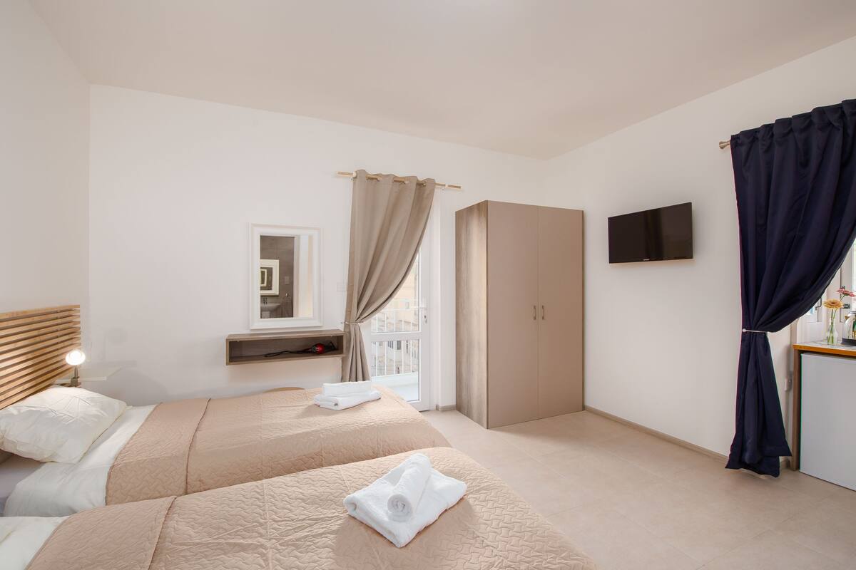 Quarto de hotel com duas camas, um guarda-roupa, uma televisão e uma janela com cortinas.