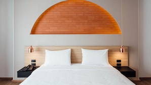 Quarto de hotel moderno com cabeceira de madeira, cama com lençóis brancos, mesa de cabeceira com telefone e duas luminárias fixadas na parede.