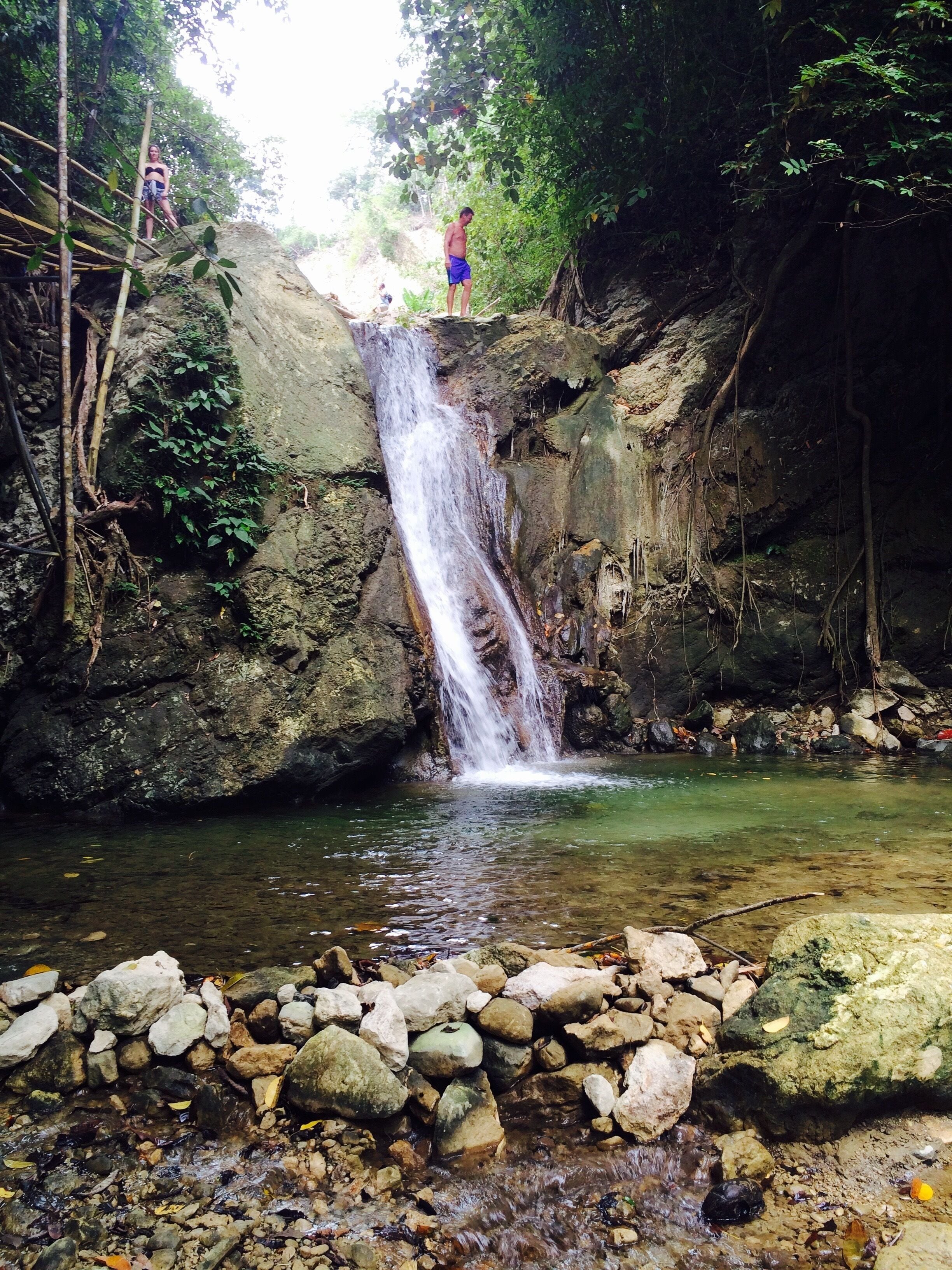 Uma cachoeira em área arborizada, com terreno rochoso e um riacho límpido na base.