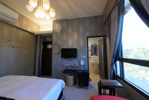 Quarto de hotel com cama, escrivaninha, televisão, janela com vista e um candelabro.