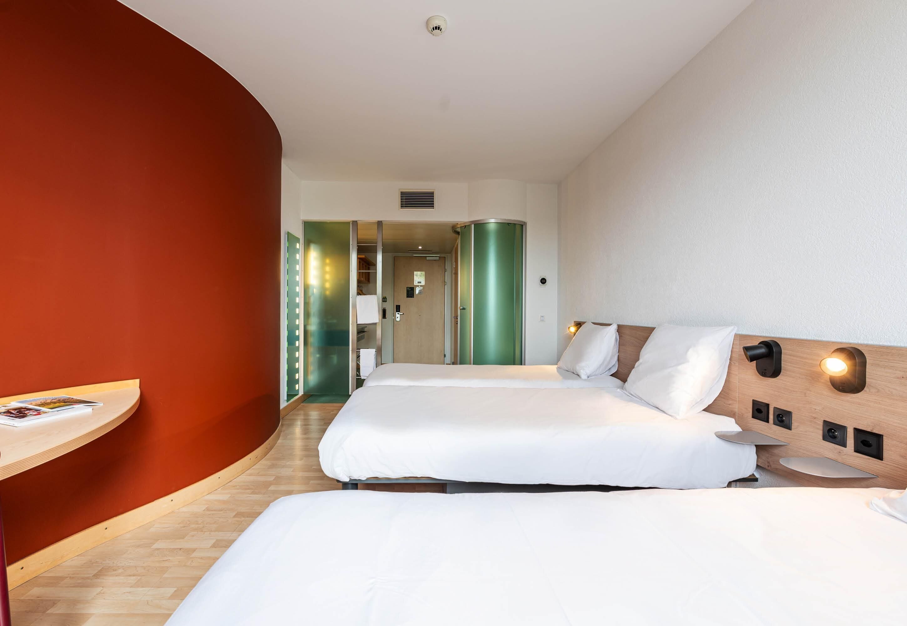 Quarto de hotel com duas camas, uma parede curva, cabeceira de madeira e uma mesinha com uma revista.
