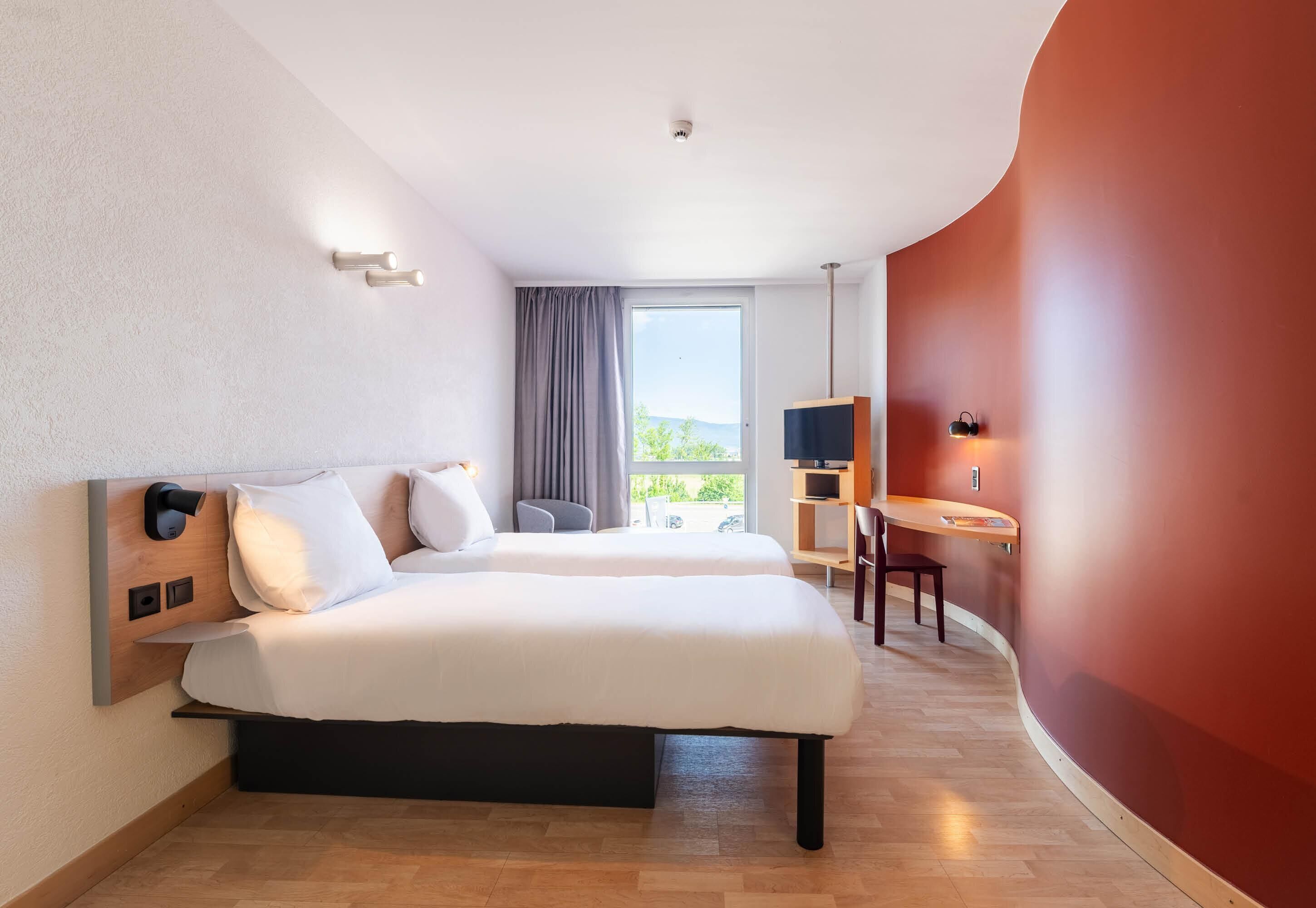 Quarto de hotel com cama, escrivaninha, cadeira, televisão e janela com vista.