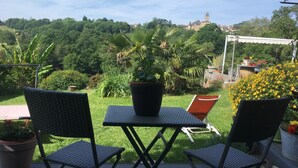 Um pátio com duas cadeiras e uma mesa, rodeado por vegetação exuberante e vista para um castelo ao longe.