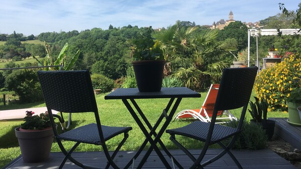 Pátio com duas cadeiras e uma mesa, rodeado por plantas em vasos e vista para uma colina com uma torre.