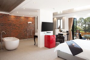 Quarto de hotel moderno com uma cama grande, televisão de tela plana, minibar vermelho, parede de tijolos e varanda com vista para árvores.