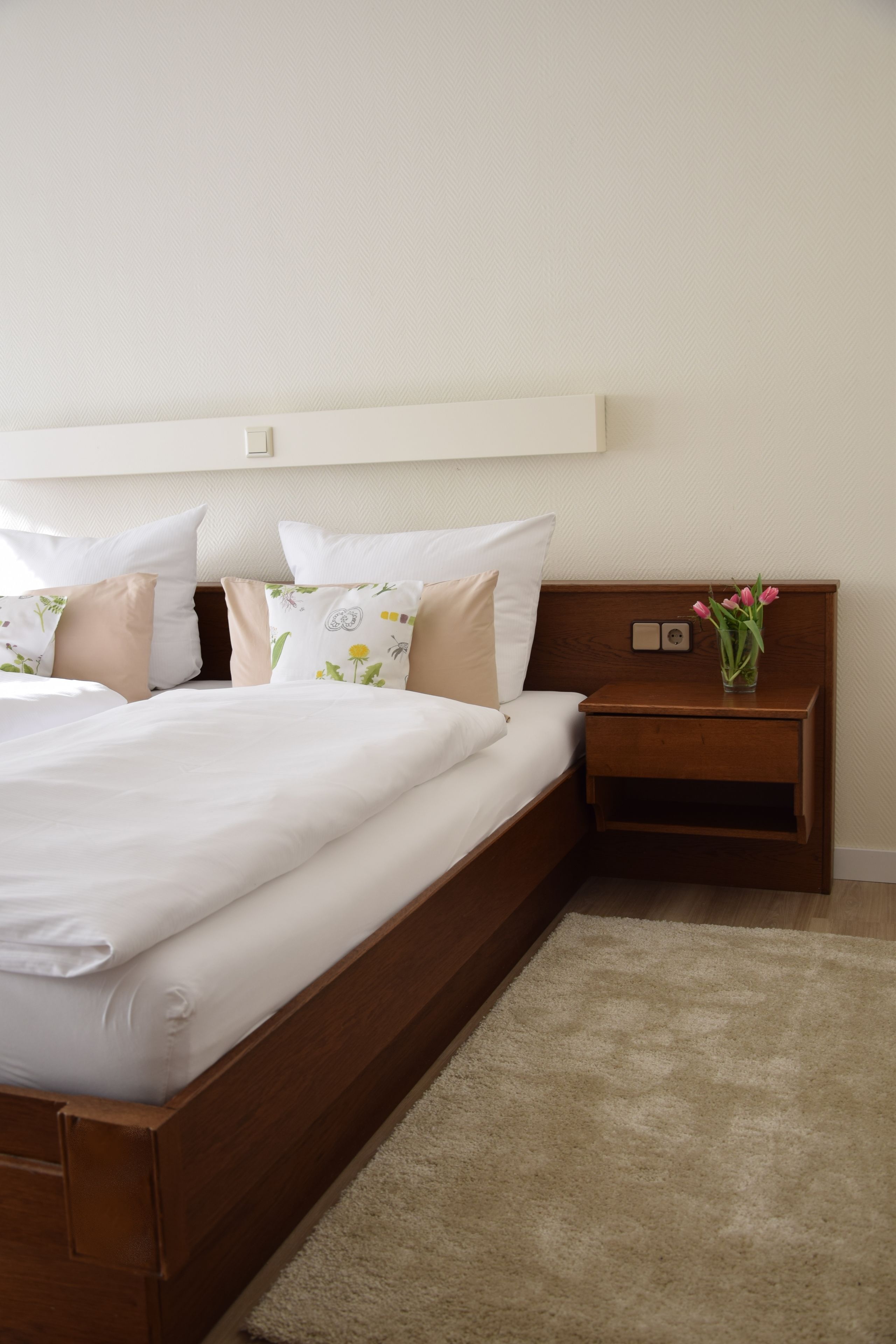 Uma cama bem arrumada com lençóis brancos e cabeceira de madeira, um criado-mudo de madeira com um vaso de flores, e um tapete bege.