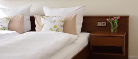 Uma cama bem arrumada com lençóis brancos e cabeceira de madeira, um criado-mudo de madeira com um vaso de flores, e um tapete bege.