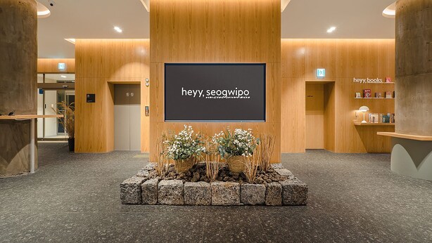 Uma área de recepção com uma tela grande exibindo "hey, seogwipo" e um pequeno arranjo de plantas.