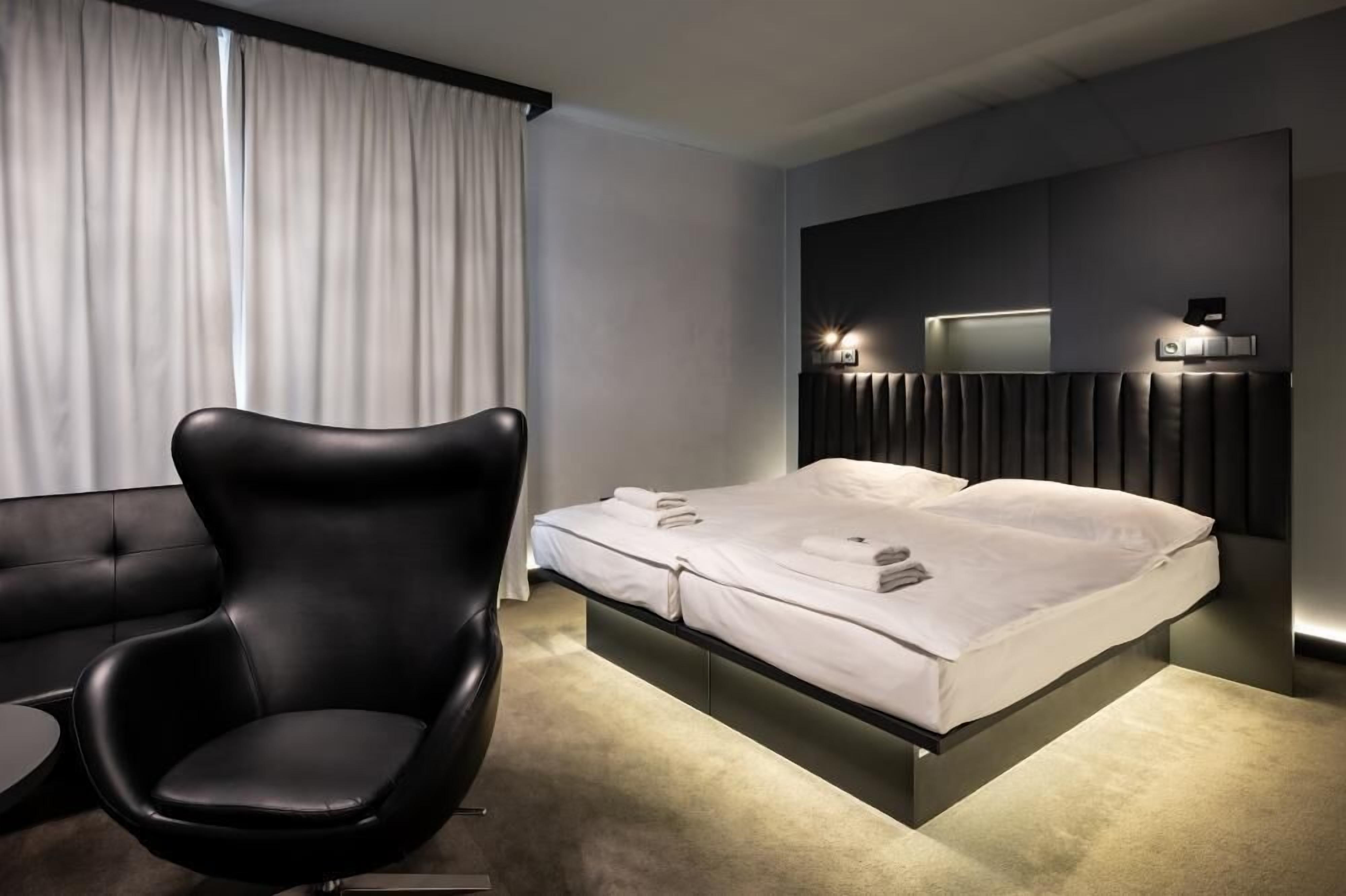 Quarto de hotel moderno com uma cama grande, uma poltrona de couro preto e um aplique na parede.