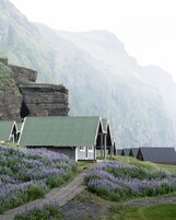 Fileira de cabanas de madeira com telhados verdes, com montanhas e um campo de flores roxas ao fundo.