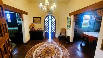 Um quarto com piso de madeira, tapete estampado, lustre, porta com detalhe em vidro decorativo e um espaço de jantar com mesa e cadeiras.