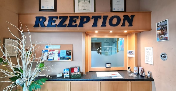 Um balcão de recepção com uma placa que diz 'RECEPTION' em cima. Há certificados emoldurados na parede, um mural de avisos com diversos papéis e uma porta de vidro com um logotipo.