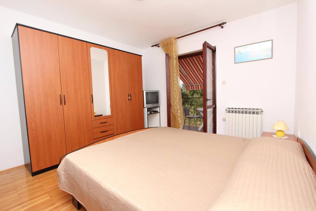 Quarto com cama grande, guarda-roupa de madeira, televisão e janela com vista para vegetação.
