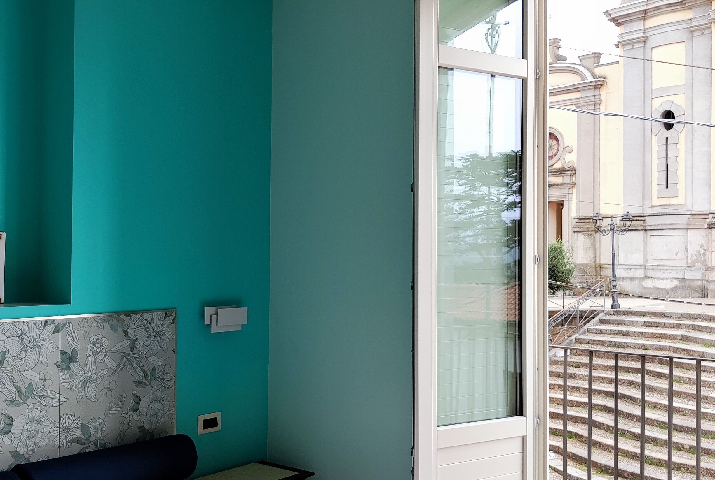 Quarto com paredes verde-água, cabeceira com padrão marmorizado e janela com vista para um edifício com escadas e corrimão.