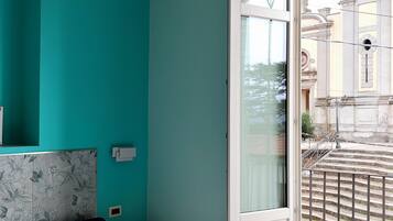 Quarto com paredes verde-água, cabeceira com padrão marmorizado e janela com vista para um edifício com escadas e corrimão.