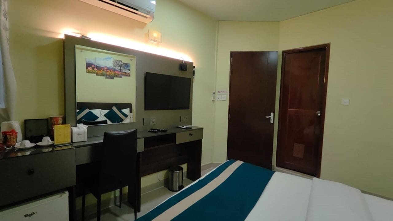Quarto de hotel com uma cama, uma escrivaninha com computador, uma cadeira, um guarda-roupa e uma pequena lixeira.
