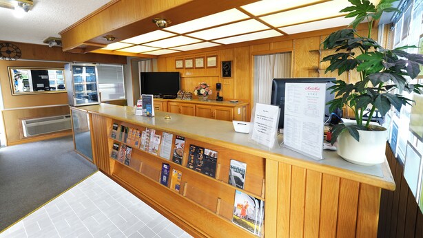Uma área de recepção com balcão de madeira, televisão, quadro de cardápio e um vaso com planta.