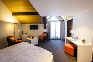 Quarto de hotel com cama, escrivaninha, cadeira, mesa pequena, televisão e janela com cortinas.