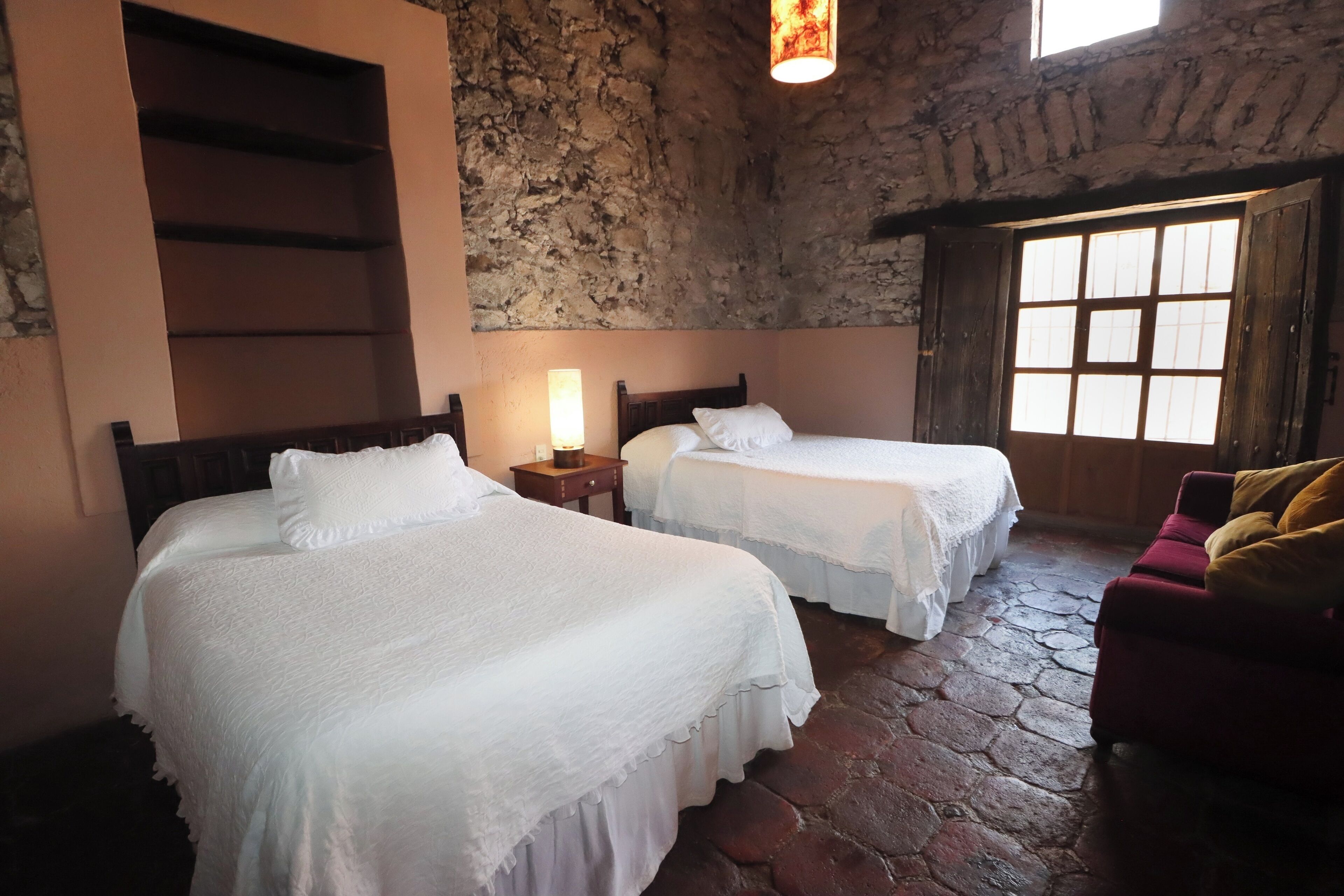 Quarto com duas camas, piso de madeira, parede de pedra e janela com persianas de madeira.
