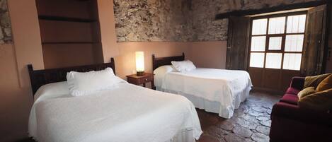 Quarto com duas camas, piso de madeira, parede de pedra e janela com persianas de madeira.