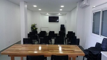 Uma sala de conferências com uma longa mesa de madeira, várias cadeiras pretas, uma televisão de tela plana grande na parede e uma janela do lado direito.