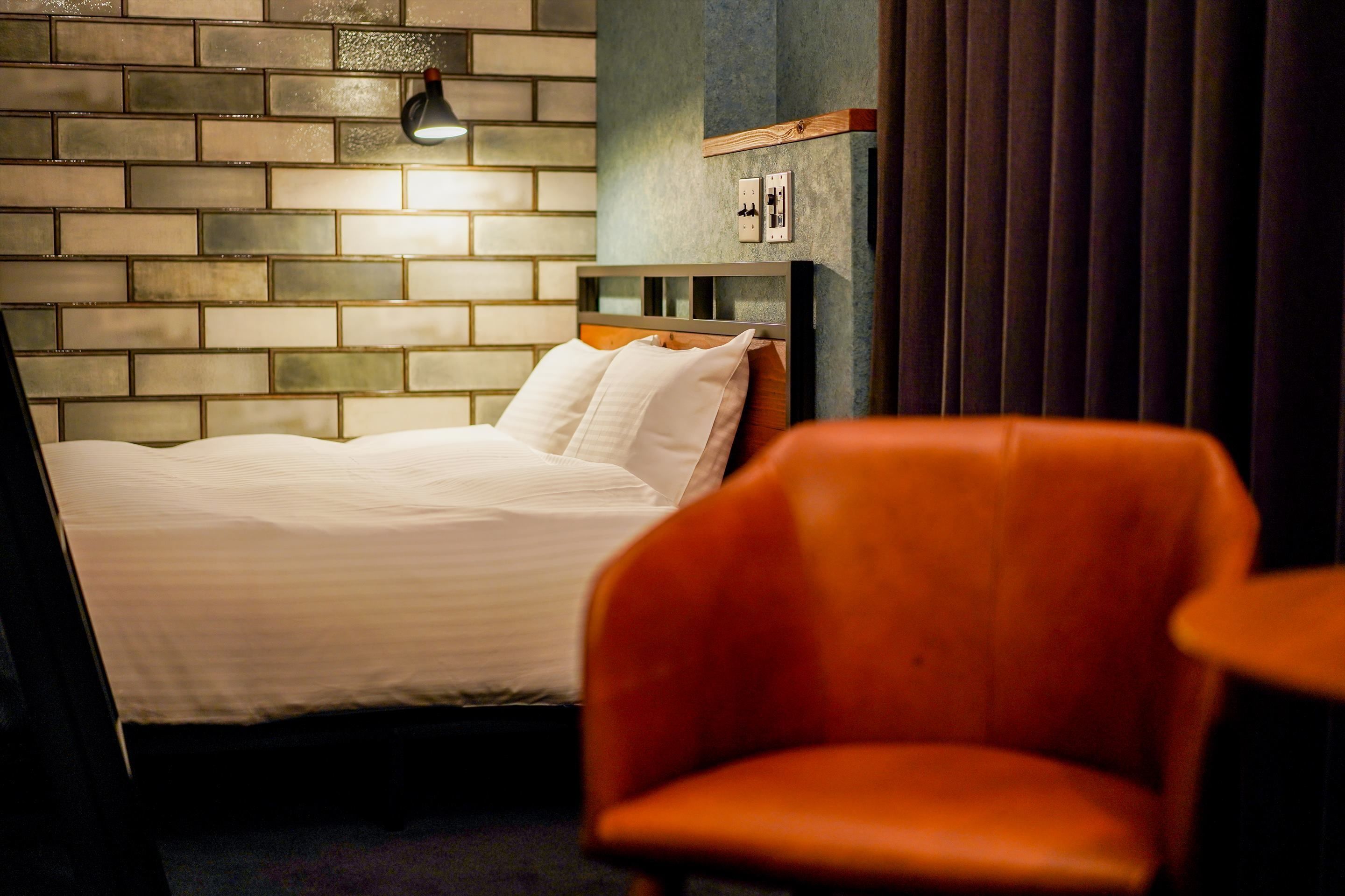 Quarto de hotel com parede em padrão tijolo, cama com roupa de cama branca, cabeceira de madeira e uma cadeira laranja.