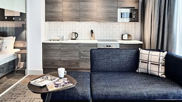 Cozinha moderna com micro-ondas embutido, máquina de café e lava-louças. Há um sofá azul com almofada xadrez e uma pequena mesa com uma revista e uma xícara de café.