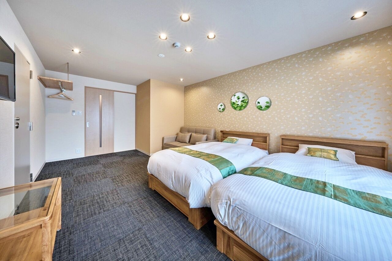 Quarto de hotel com duas camas, cabeceira de madeira, piso coberto por carpete, mesa de vidro e televisão fixada na parede.