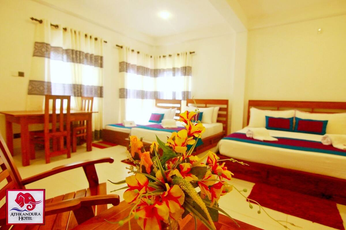 Quarto de hotel com duas camas, uma cadeira de madeira, uma escrivaninha e um vaso de flores.