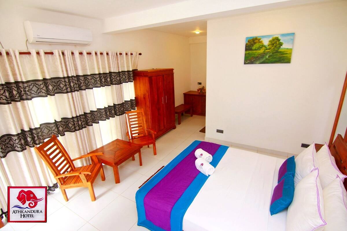 Quarto de hotel com cama, uma cadeira de madeira, uma mesinha, escrivaninha e um quadro na parede.
