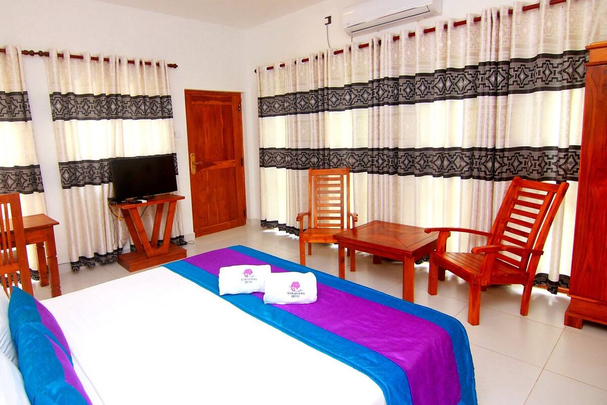 Quarto de hotel com cama, televisão, escrivaninha e duas cadeiras.