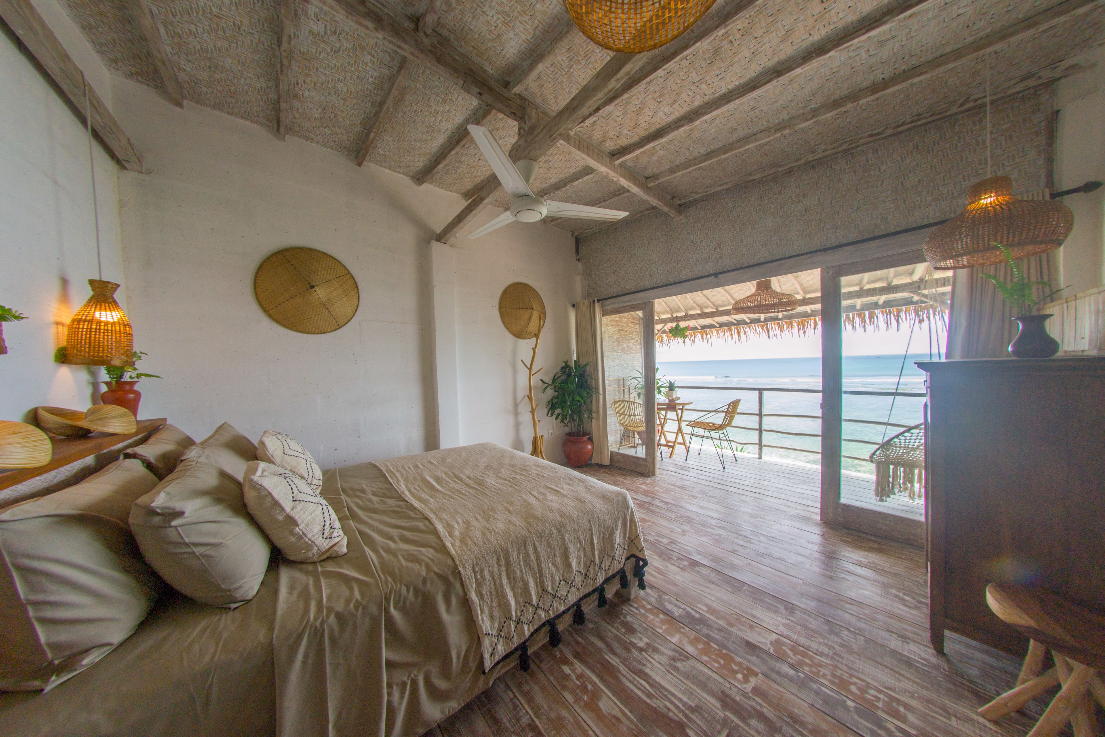 Quarto com cama, ventilador de teto, janela ampla e vista para o oceano.