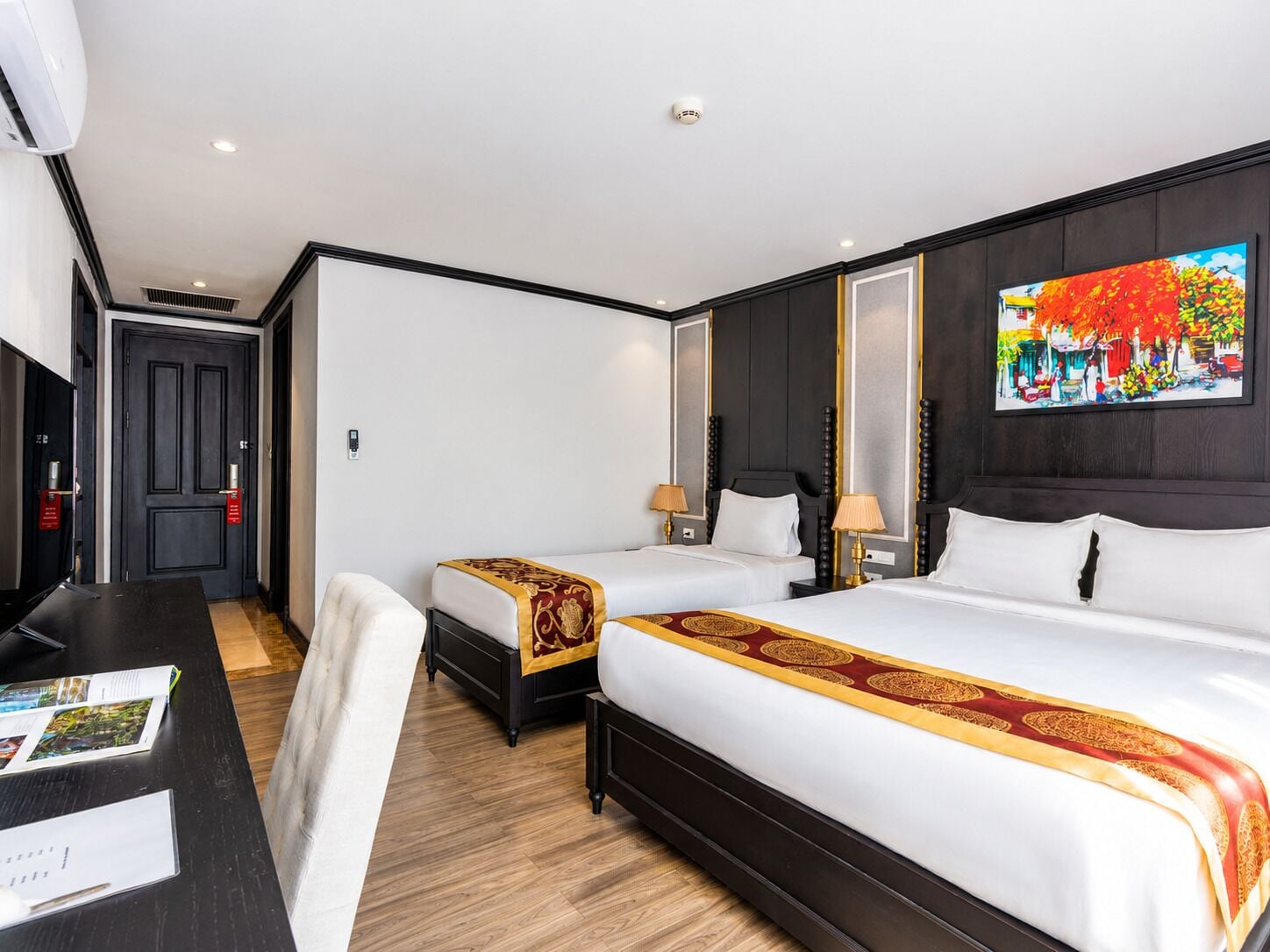 Quarto de hotel com duas camas, uma escrivaninha, uma cadeira, uma TV e um quadro na parede.