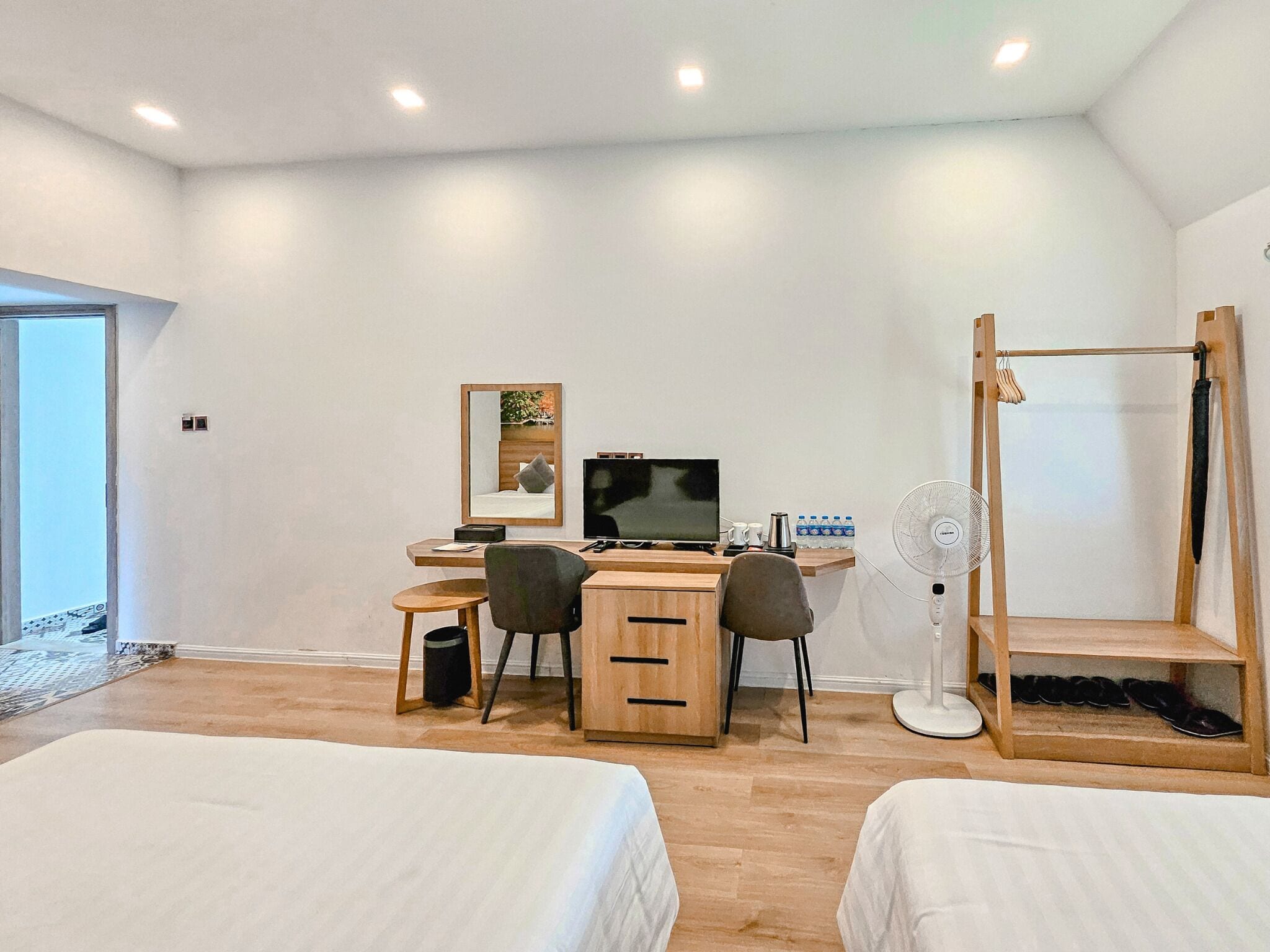Quarto moderno com duas camas, uma escrivaninha com computador, uma cadeira, um ventilador de chão e uma estante de madeira.