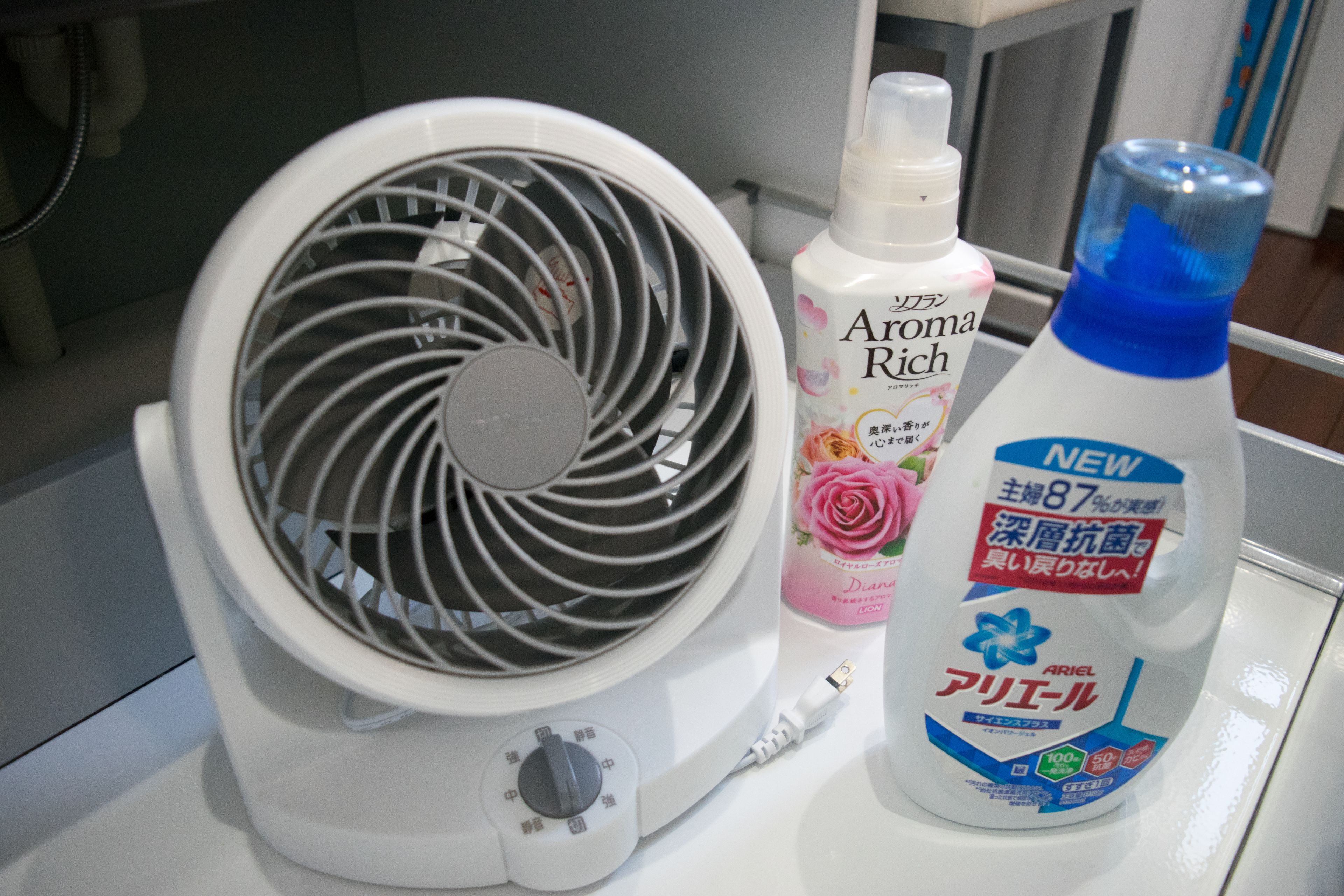 Um ventilador branco, uma garrafa de Aroma Rich e uma garrafa de detergente Ariel sobre uma superfície branca.
