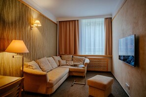 Quarto de hotel moderno com sofá modular, uma mesa pequena, luminária, radiador, televisão e janela com cortinas.