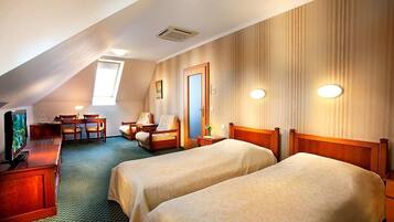 Quarto de hotel com duas camas, uma secretária com televisão, uma cadeira, uma pequena mesa com um vaso e uma janela que oferece luz natural.