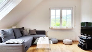Uma sala de estar moderna com um sofá cinzento, uma mesa de centro de vidro, uma televisão e uma janela com vista para o verde.