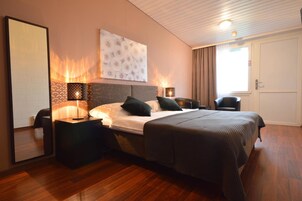 Quarto de hotel com uma cama grande, duas mesas de cabeceira, uma cadeira, uma janela com cortinas e uma porta.