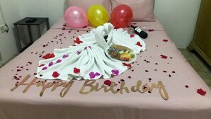 Uma cama com colcha rosa, balões, um buquê de flores e uma placa com os dizeres 'Feliz Aniversário'.