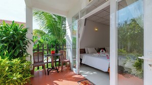 Quarto com cama, uma pequena mesa e cadeiras, varanda com plantas e porta de vidro deslizante para o exterior.