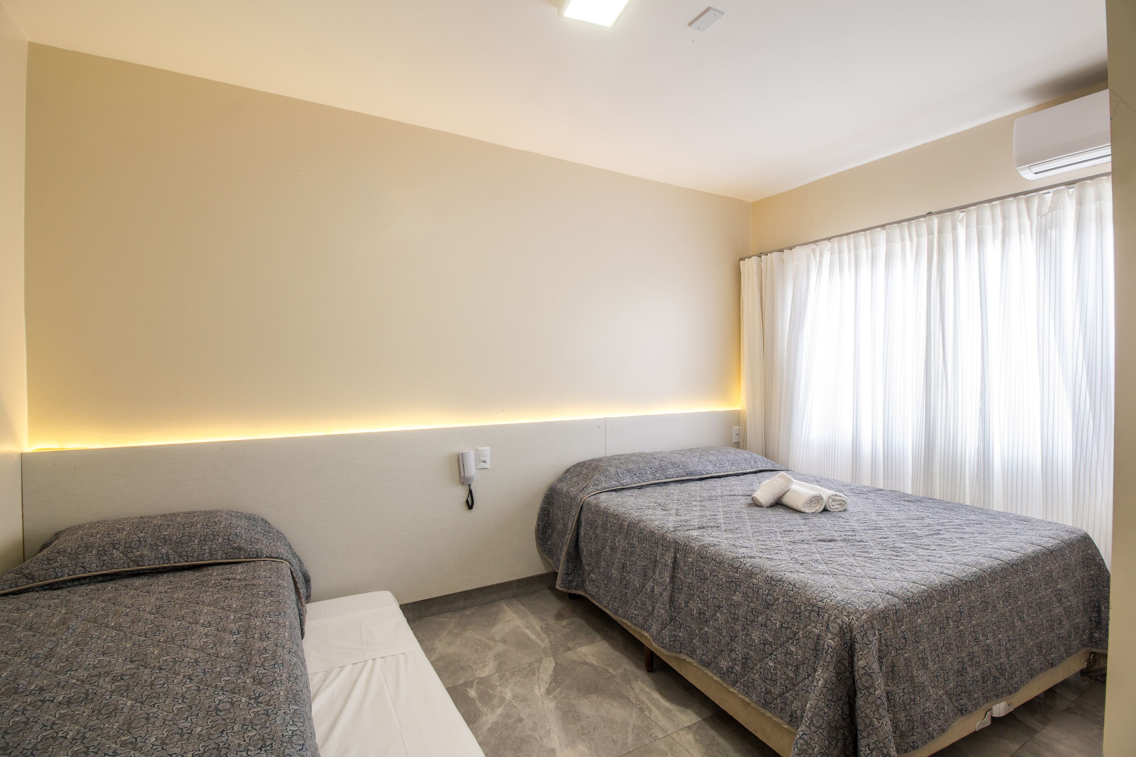 Quarto de hotel com duas camas de solteiro, mesa de cabeceira, telefone fixado na parede e janela com cortinas.