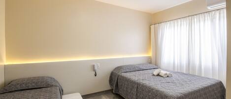 Quarto de hotel com duas camas de solteiro, mesa de cabeceira, telefone fixado na parede e janela com cortinas.