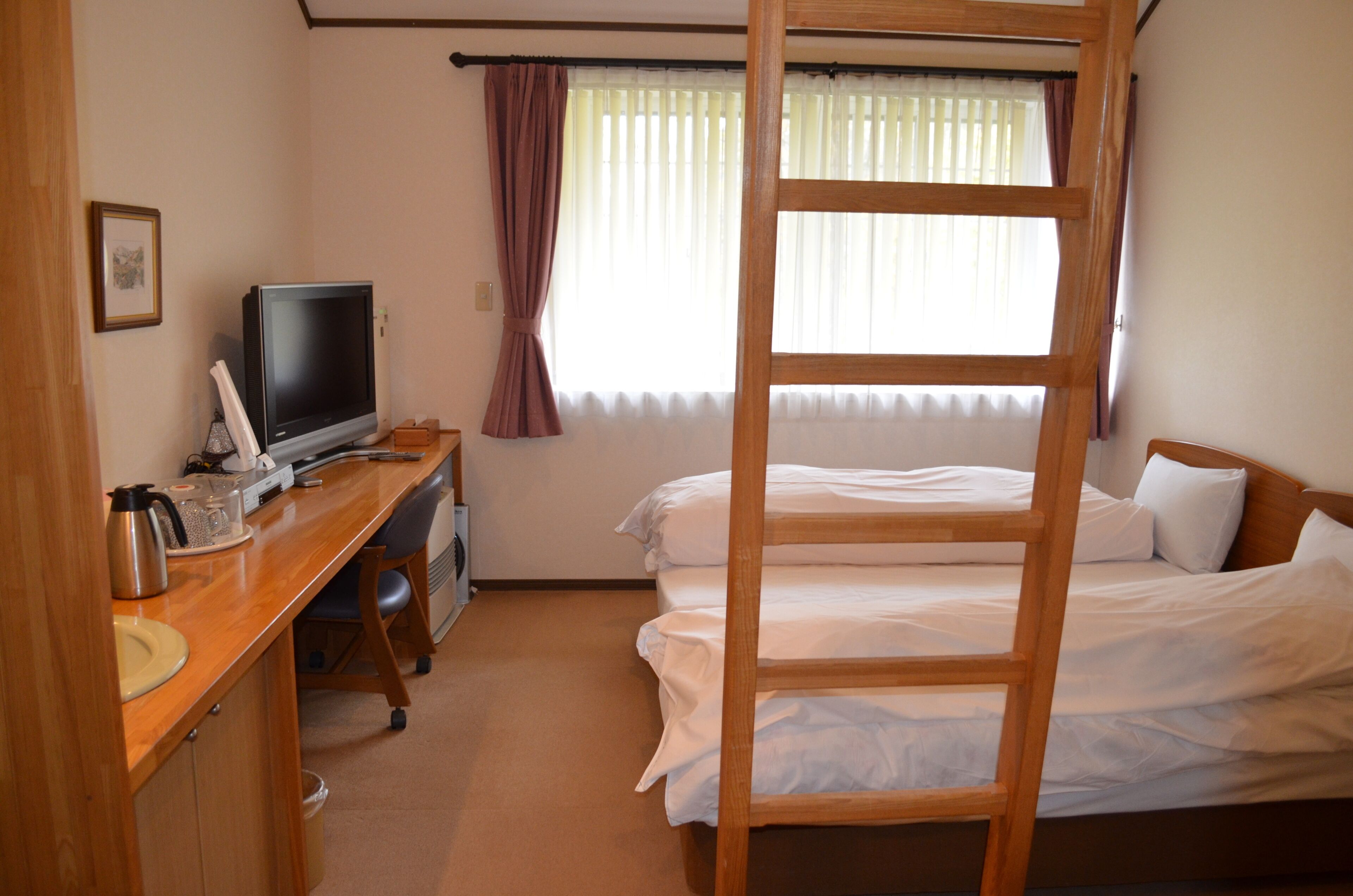 Quarto com beliche, escrivaninha com televisão, cadeira, mesa pequena com chaleira e janela com cortinas.